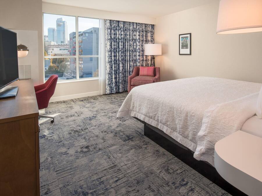Quarto de hotel com uma cama grande, uma escrivaninha com televisão, uma cadeira vermelha, uma janela com cortinas e vista para a cidade.
