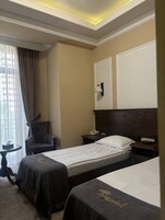Quarto de hotel com duas camas, uma cadeira, mesa de cabeceira com abajur e janela com cortinas.