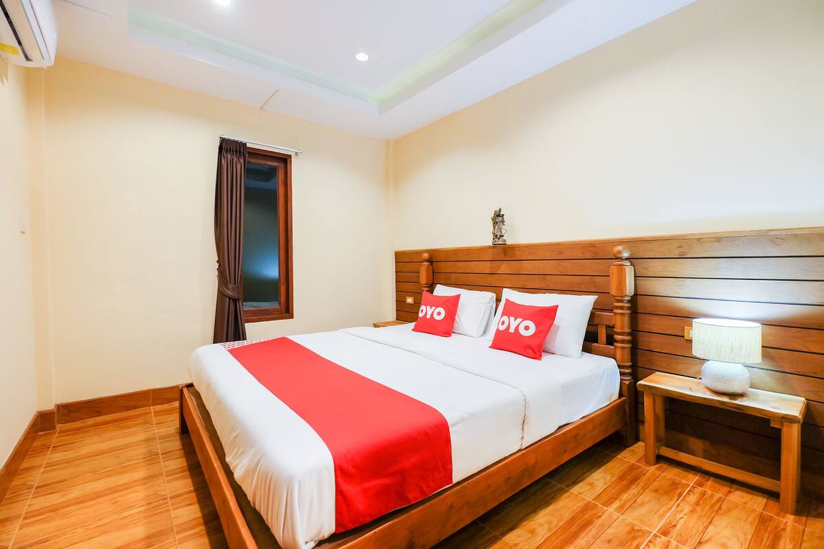 Quarto de hotel com cama de madeira, mesa de cabeceira com abajur e janela com cortinas.