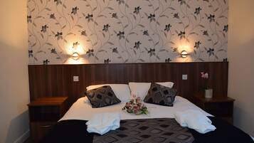 Quarto de hotel com cama, duas mesas de cabeceira, papel de parede floral e duas luminárias de parede.