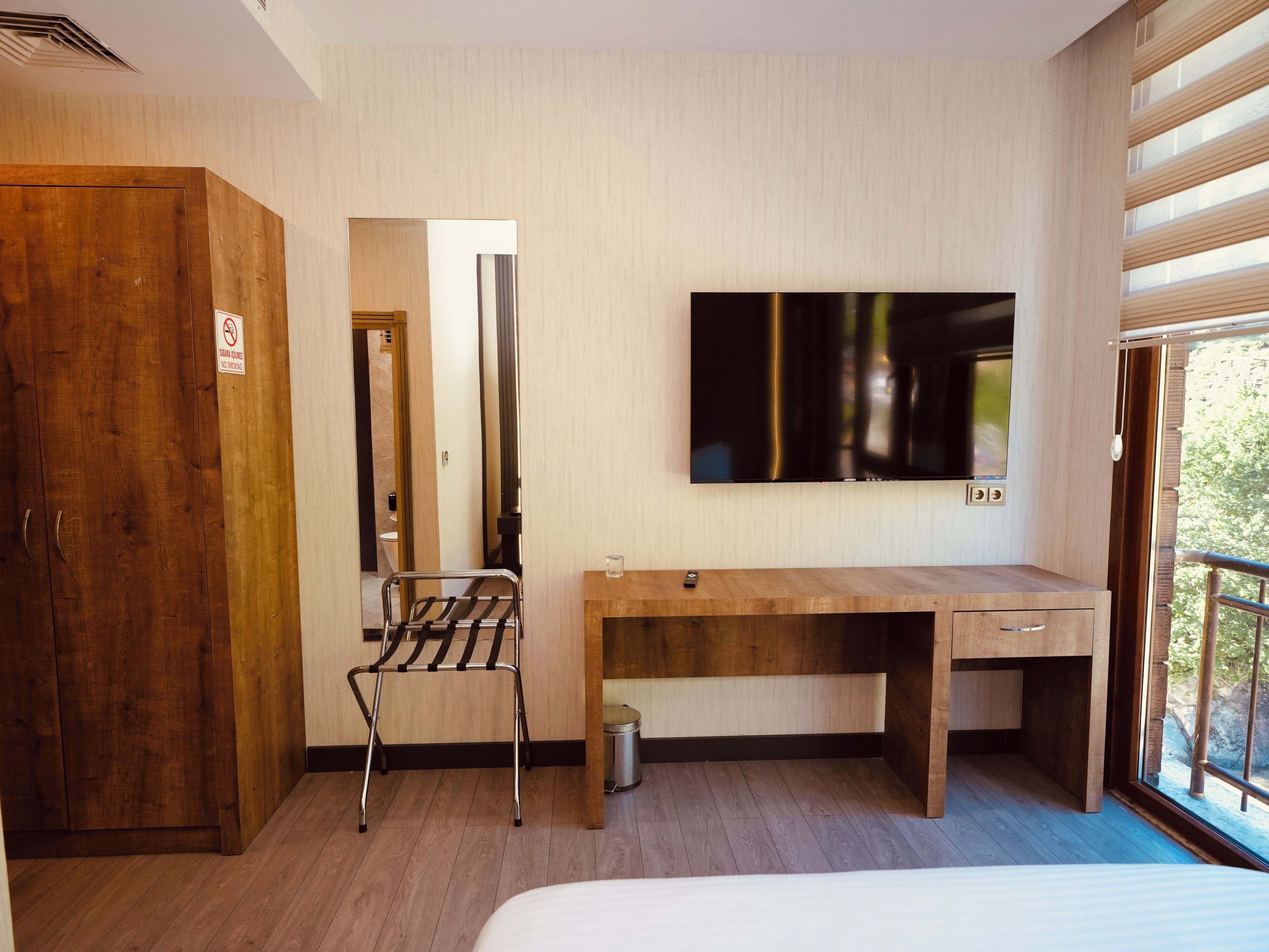 Quarto de hotel com guarda-roupa de madeira, uma escrivaninha com televisão, uma cadeira e janela com vista.