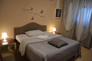 Uma cama bem arrumada com um cobre-leito cinza estampado, duas almofadas e uma toalha dobrada. Duas luminárias de cabeceira estão sobre mesas de cabeceira de madeira. O quarto tem um tapete bege e uma parede com adesivos de borboletas e uma moldura com um quadro.
