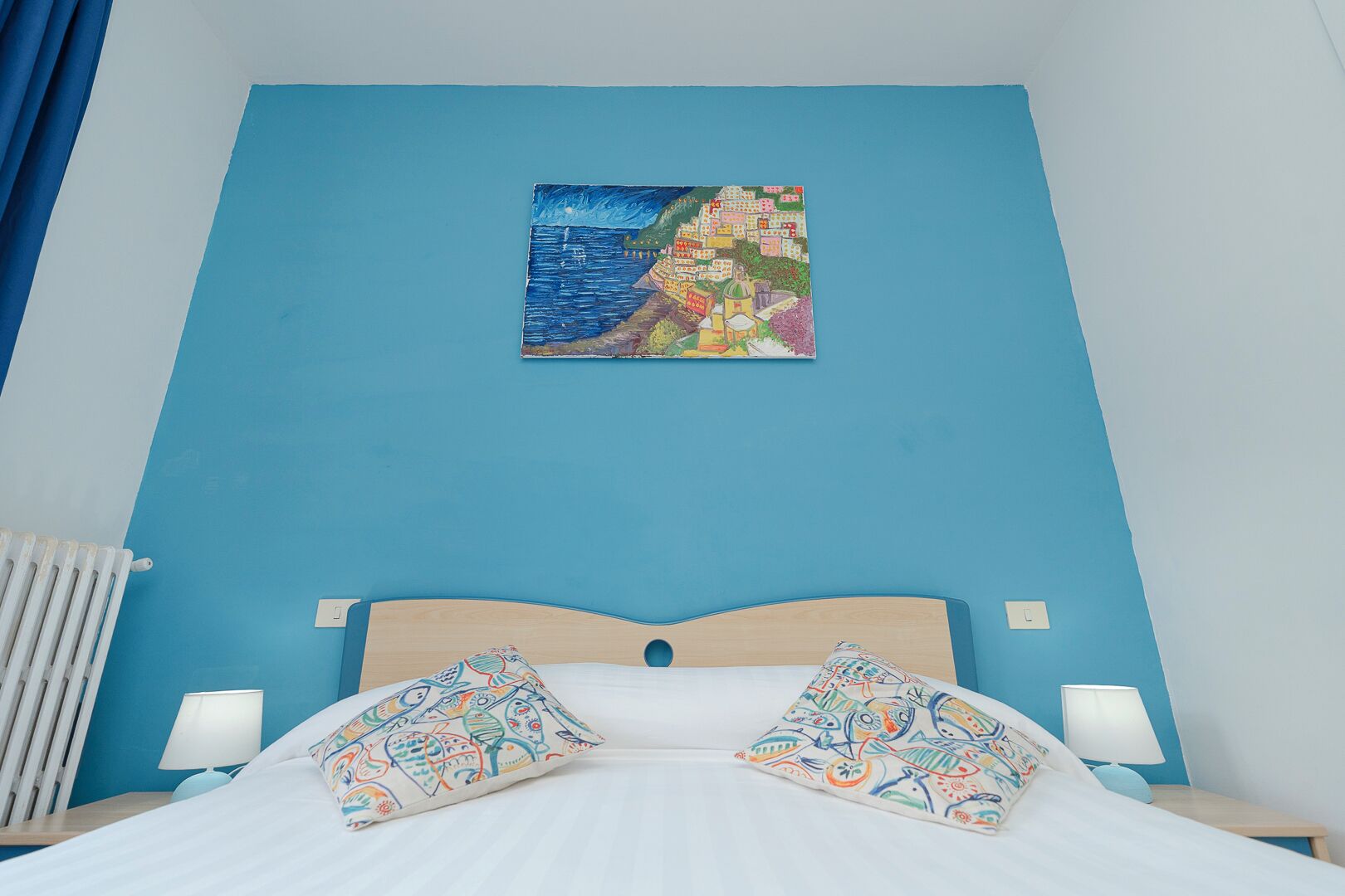 Quarto com cama, duas almofadas, mesa de cabeceira, abajur, radiador e um quadro na parede.