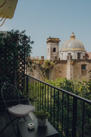 Varanda com cadeira, uma pequena mesa e um vaso de planta. Ao fundo, um edifício com cúpula e campanário.