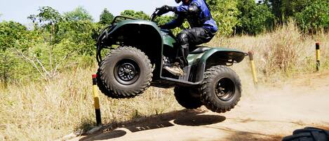 Uma pessoa conduz uma moto de trilha por um caminho de terra, rodeada por vegetação.