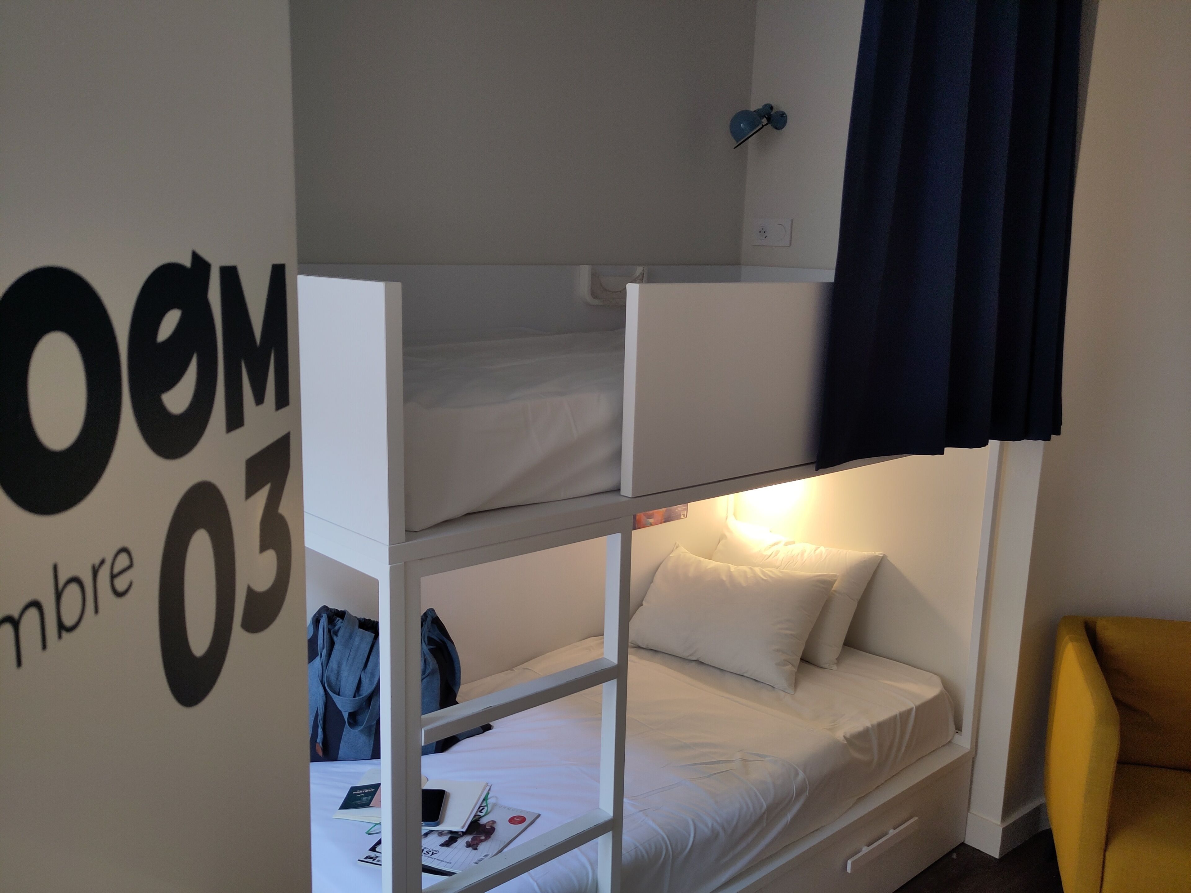 Quarto com beliche, luminária na parede, mala e uma cadeira amarela.