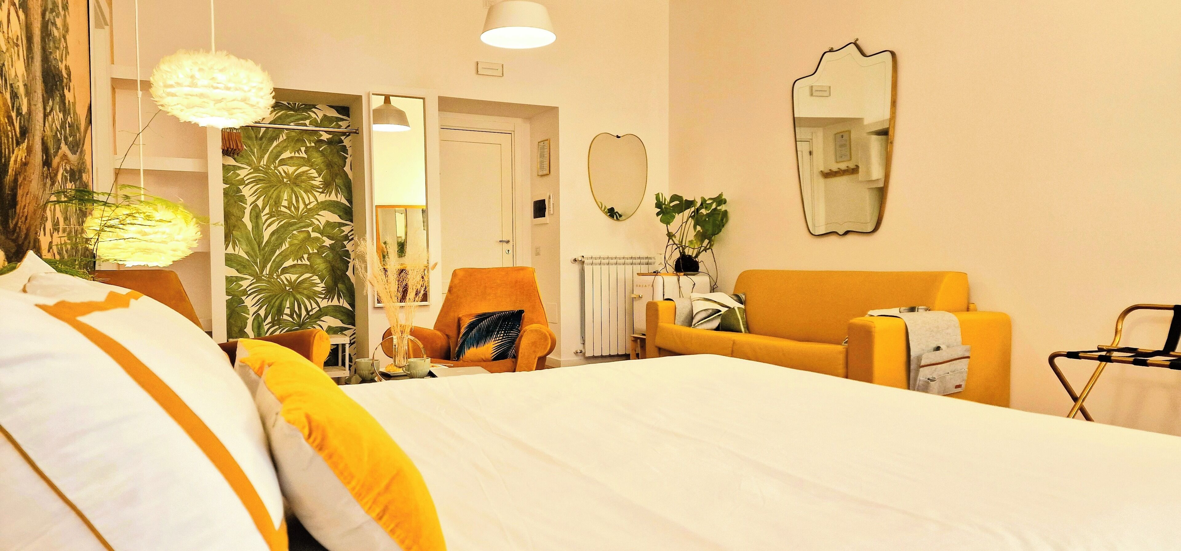 Quarto de hotel com cama, poltrona amarela, sofá, mesinha, espelho e planta em vaso.