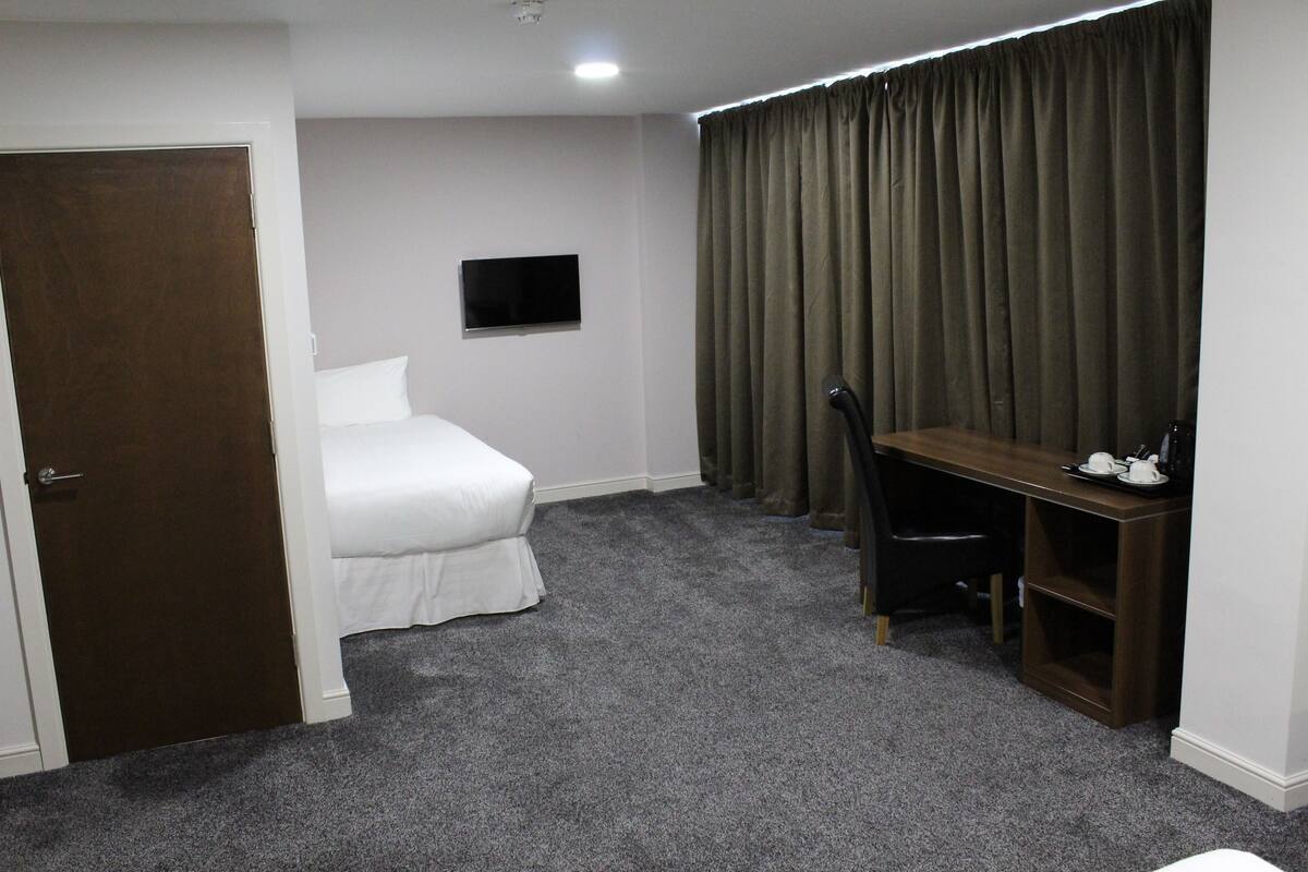Quarto de hotel com cama, escrivaninha, cadeira, televisão e porta.