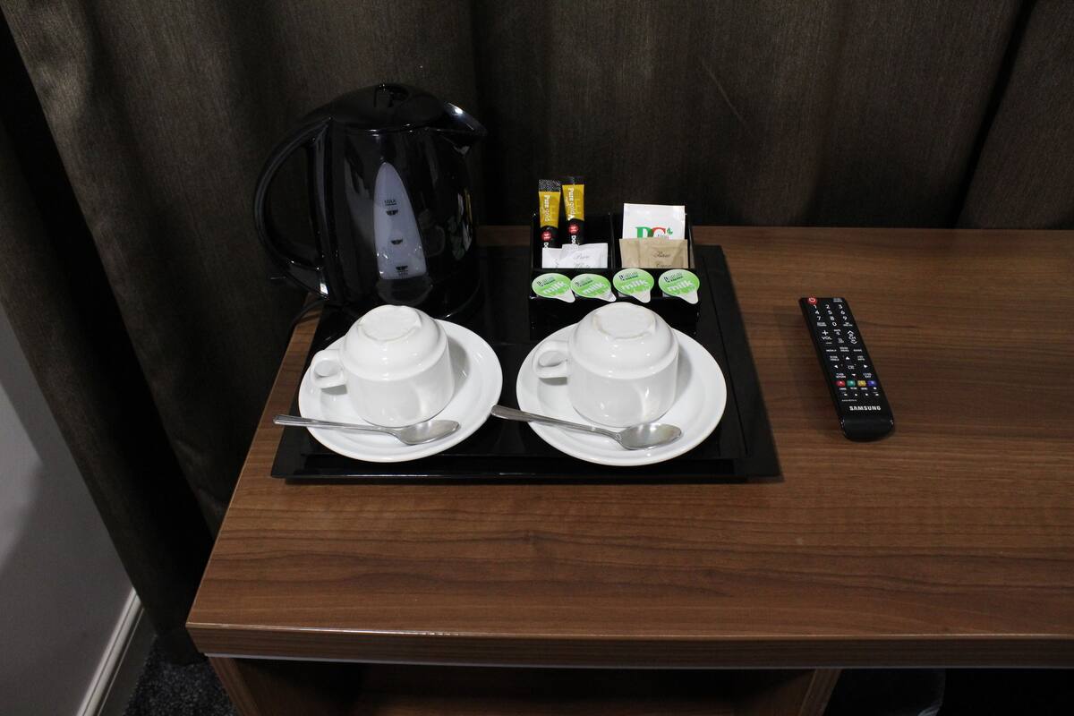 Quarto de hotel com uma mesa de madeira posta para o café da manhã, incluindo duas xícaras brancas, colheres, uma cafeteira preta, sachês de chá, um controle remoto e um cartão.