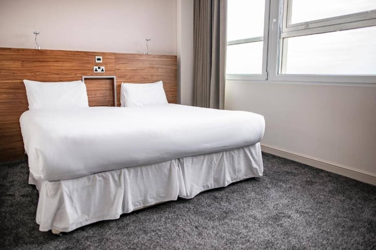 Quarto de hotel com cabeceira de madeira, uma cama grande com roupa de cama branca, um carpete cinza e uma janela com cortinas.