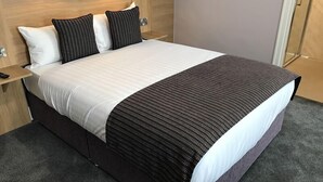 Cama bem arrumada com colcha branca e marrom escura, duas almofadas brancas e cabeceira de madeira com luzes fixadas na parede.