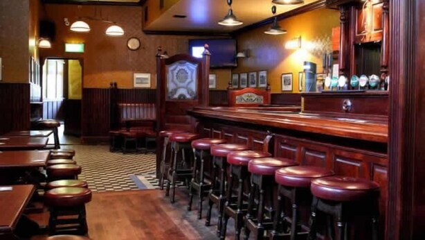 Uma área de bar com banquetas e mesas de madeira, chão xadrez e um balcão com espelho e iluminação.