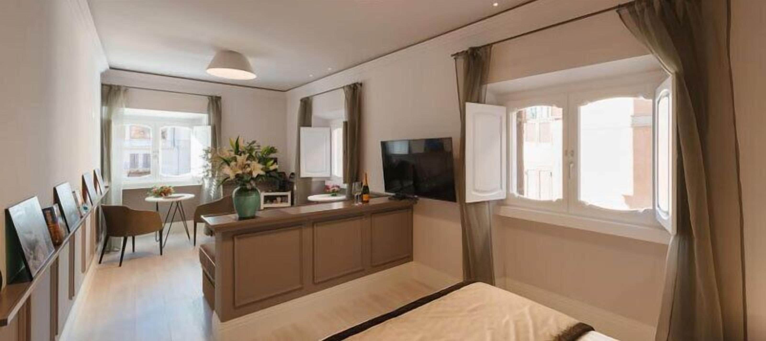 Quarto de hotel moderno com uma cama grande, uma televisão, área de refeições com mesa e cadeiras e vista da cidade através de amplas janelas.