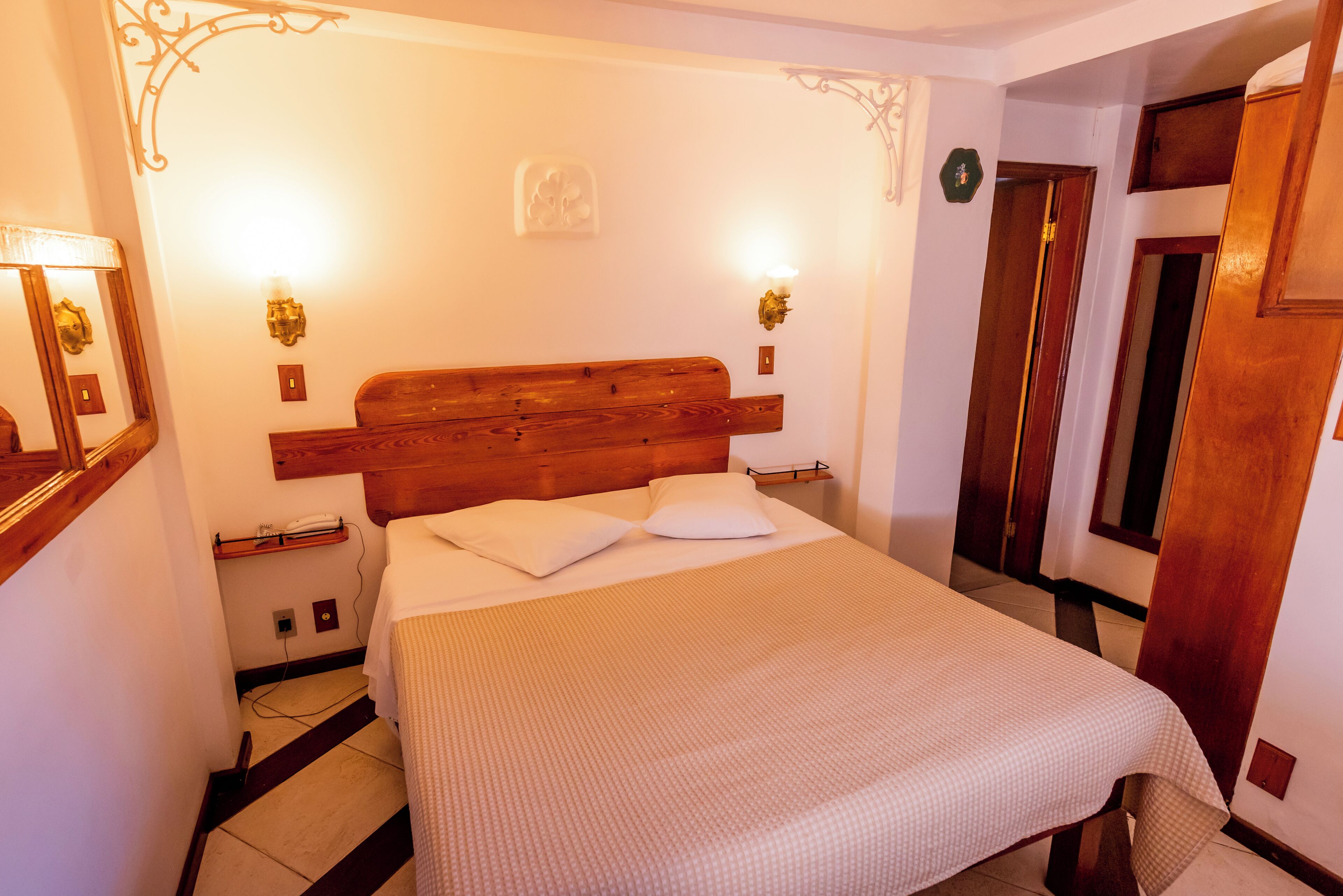 Cama bem arrumada com cabeceira de madeira, duas luminárias de cabeceira e uma pequena prateleira com um telefone.