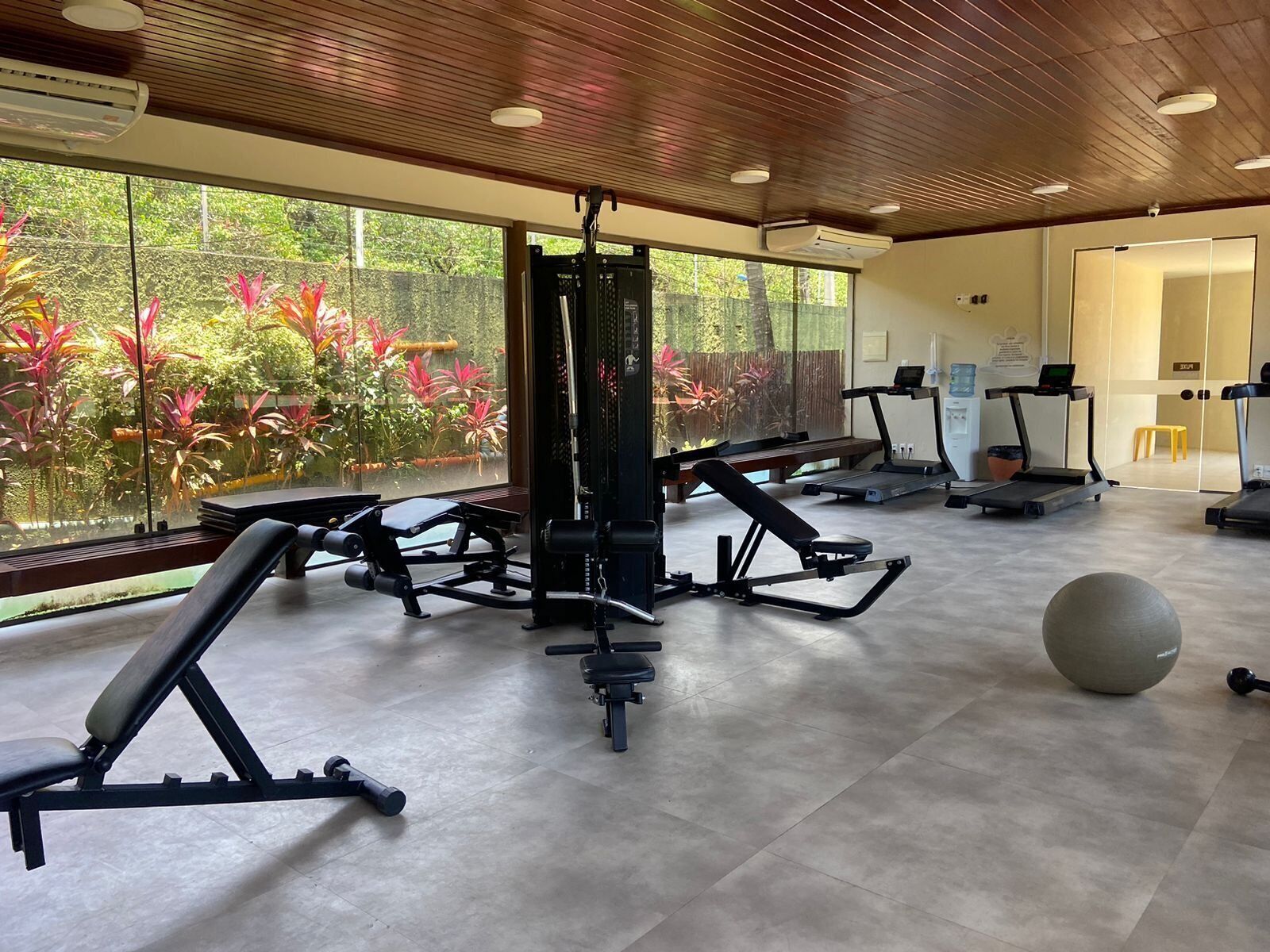 Academia com equipamentos de ginástica, uma bola de exercícios grande e vista para o verde através de amplas janelas.