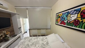 Quarto com cama, televisão, escrivaninha com um vaso de flores e um quadro abstrato colorido na parede.