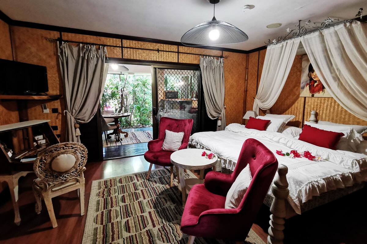 Quarto de hotel com uma cama, duas cadeiras vermelhas, uma mesa pequena, uma televisão e vista para o exterior.