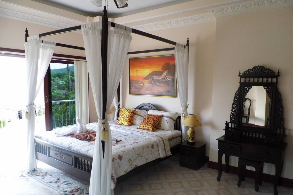 Uma cama com dossel e cortina, uma pintura de um pôr do sol, um aparador de madeira e um espelho.