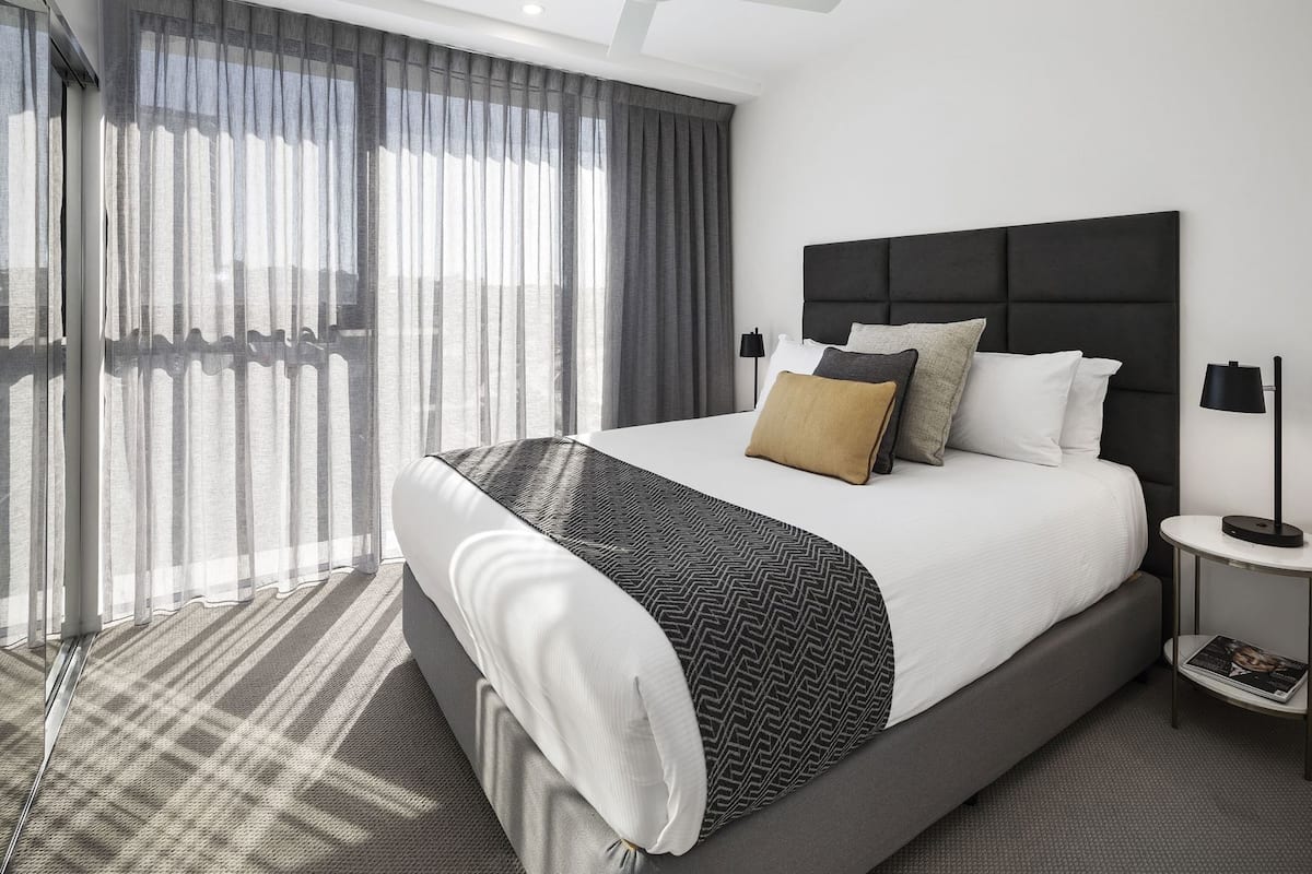 Quarto de hotel moderno com uma cama grande, uma mesinha de cabeceira e vista através de cortinas leves.