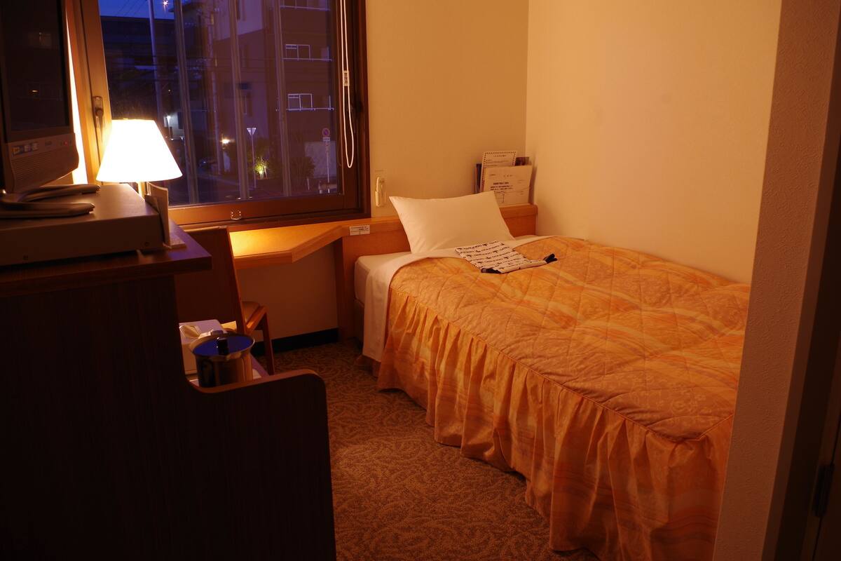 Quarto de hotel com uma cama, uma mesa com abajur, uma janela e vista da cidade à noite.