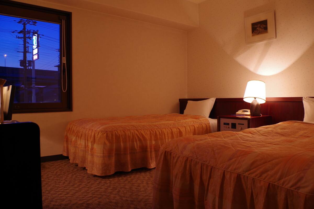 Quarto de hotel com duas camas de solteiro, uma mesa de cabeceira com abajur, uma pequena mesa e uma janela com vista para o exterior.
