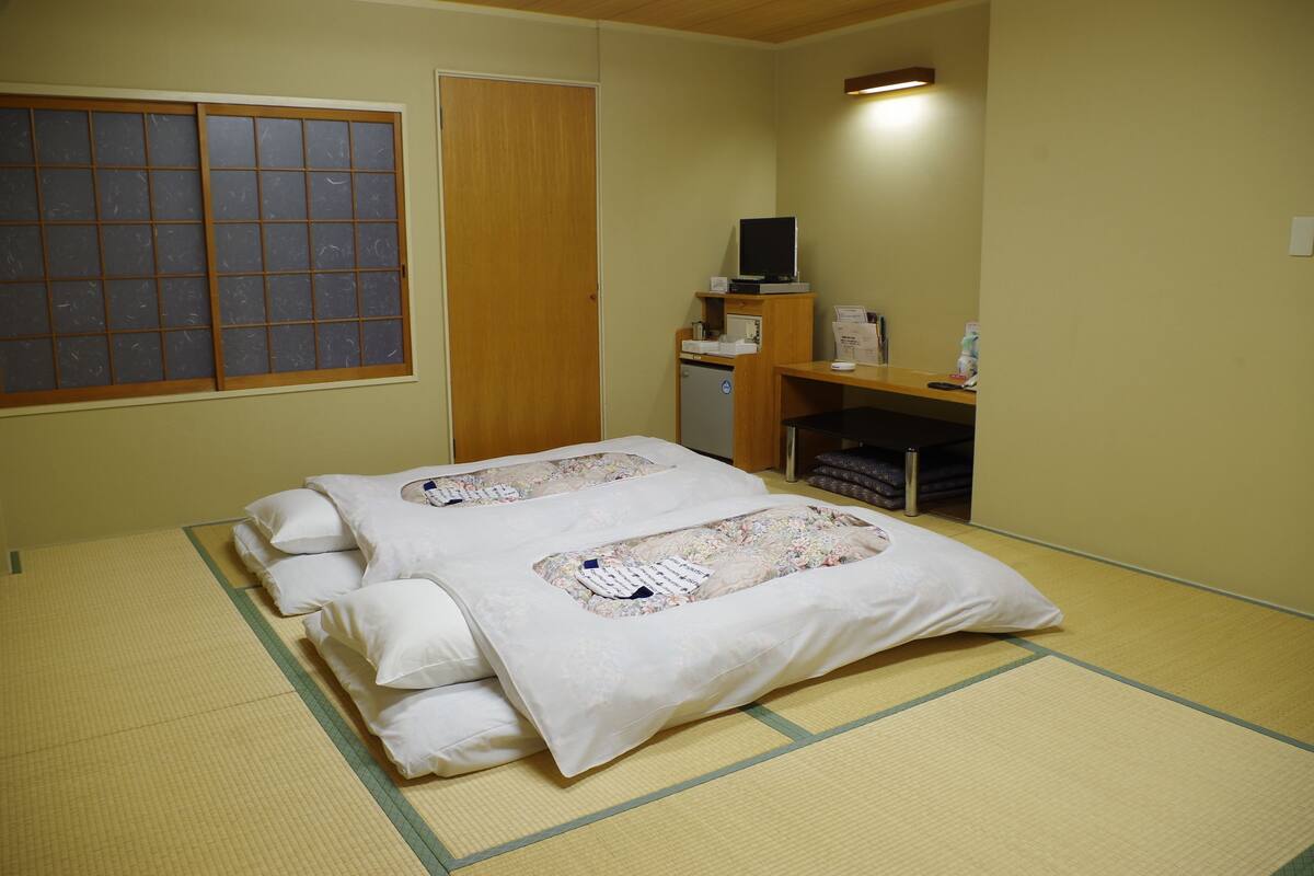 Um quarto japonês tradicional com tatames, futon, porta deslizante, televisão e uma pequena mesa com cadeira.