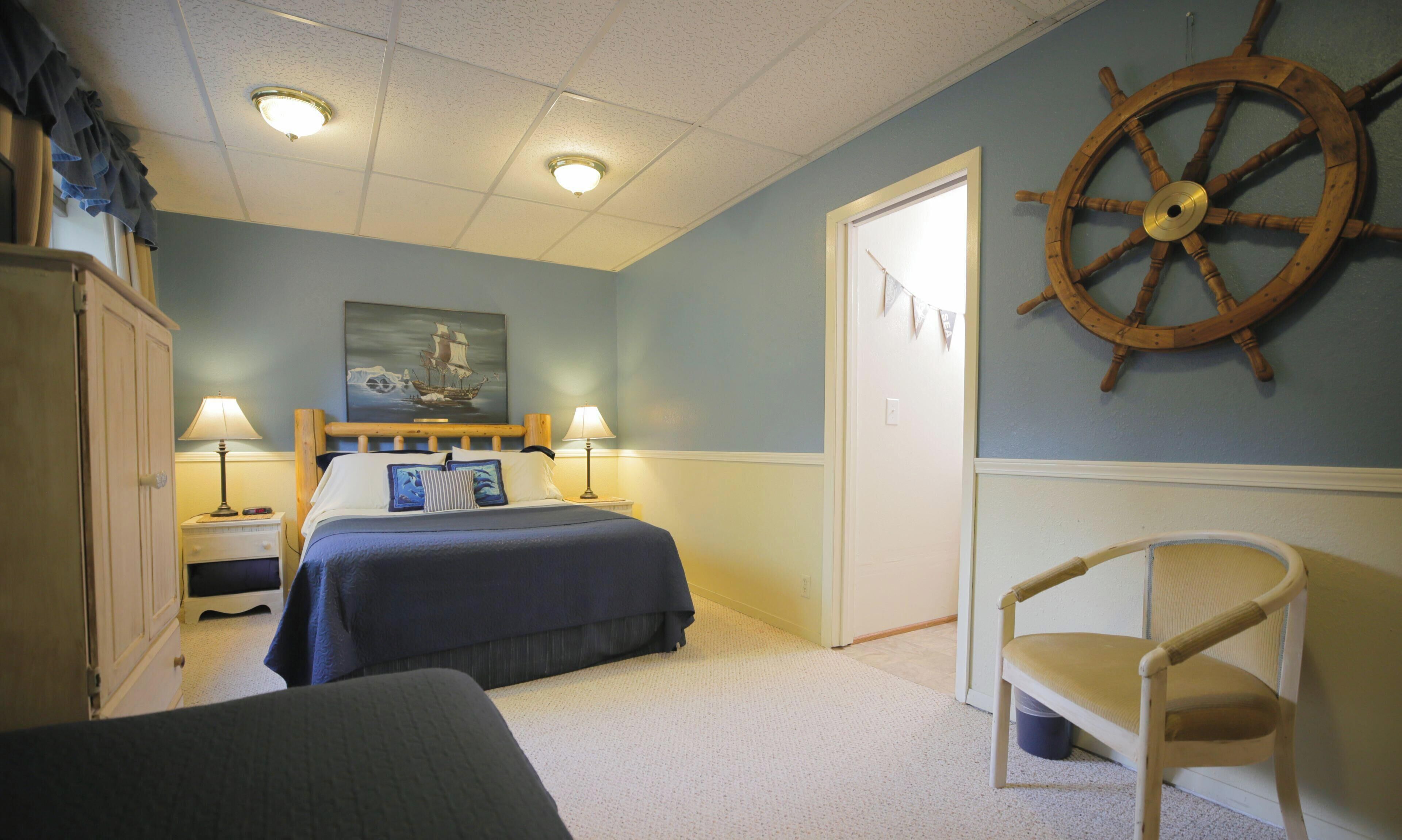 Um quarto com cama, duas luminárias de cabeceira, um leme fixado na parede e uma porta que dá para outro cômodo.