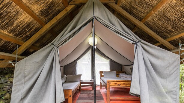 Alojamento em tenda com duas camas, uma mesa central e telhado de palha.