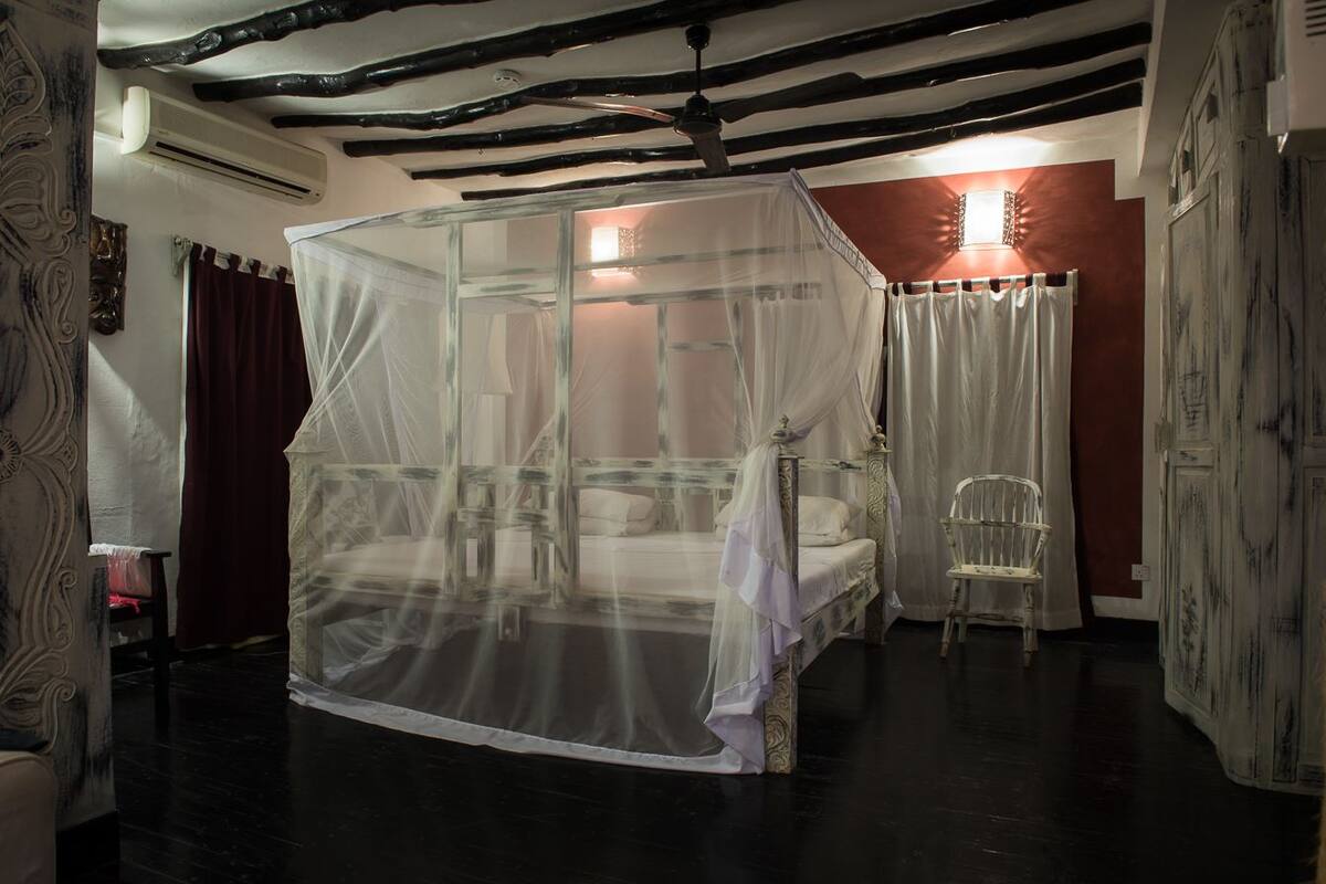 Quarto com cama de dossel coberta por um mosquiteiro, uma cadeira e ar condicionado fixado na parede.