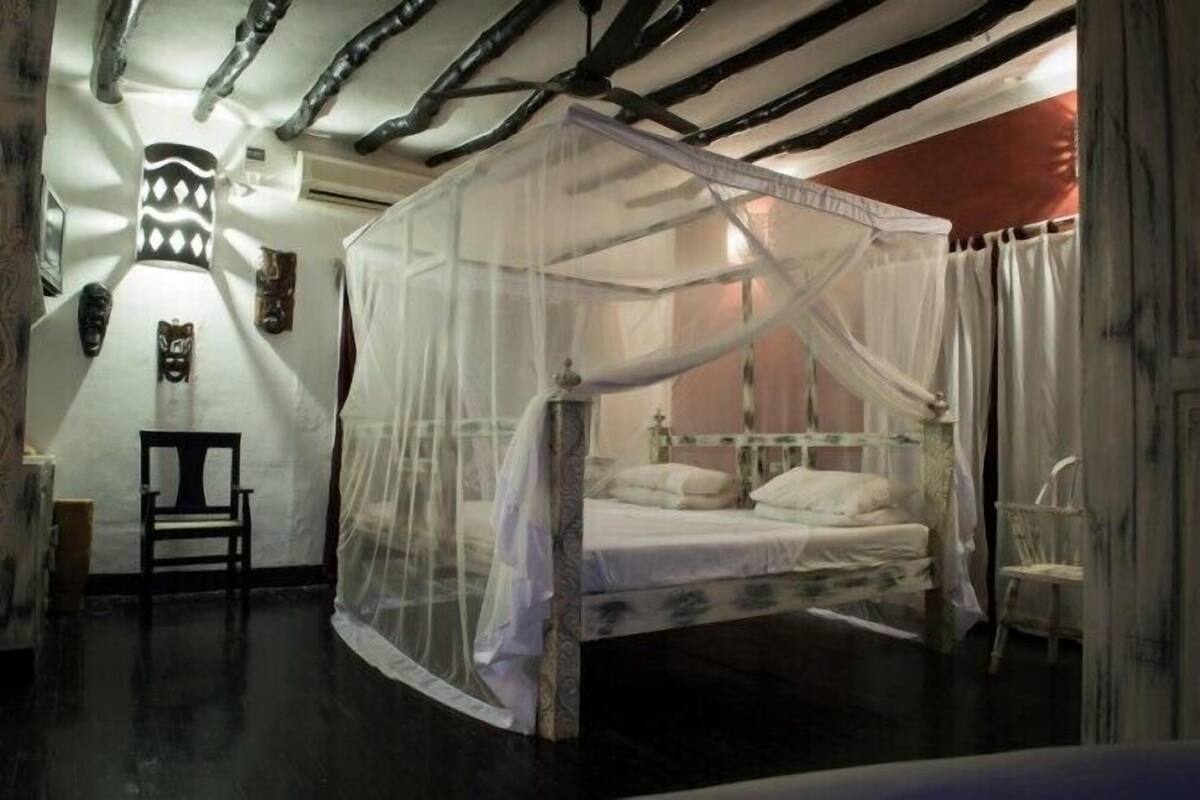 Quarto com cama sob um dossel branco, uma cadeira e parede com máscaras decorativas.