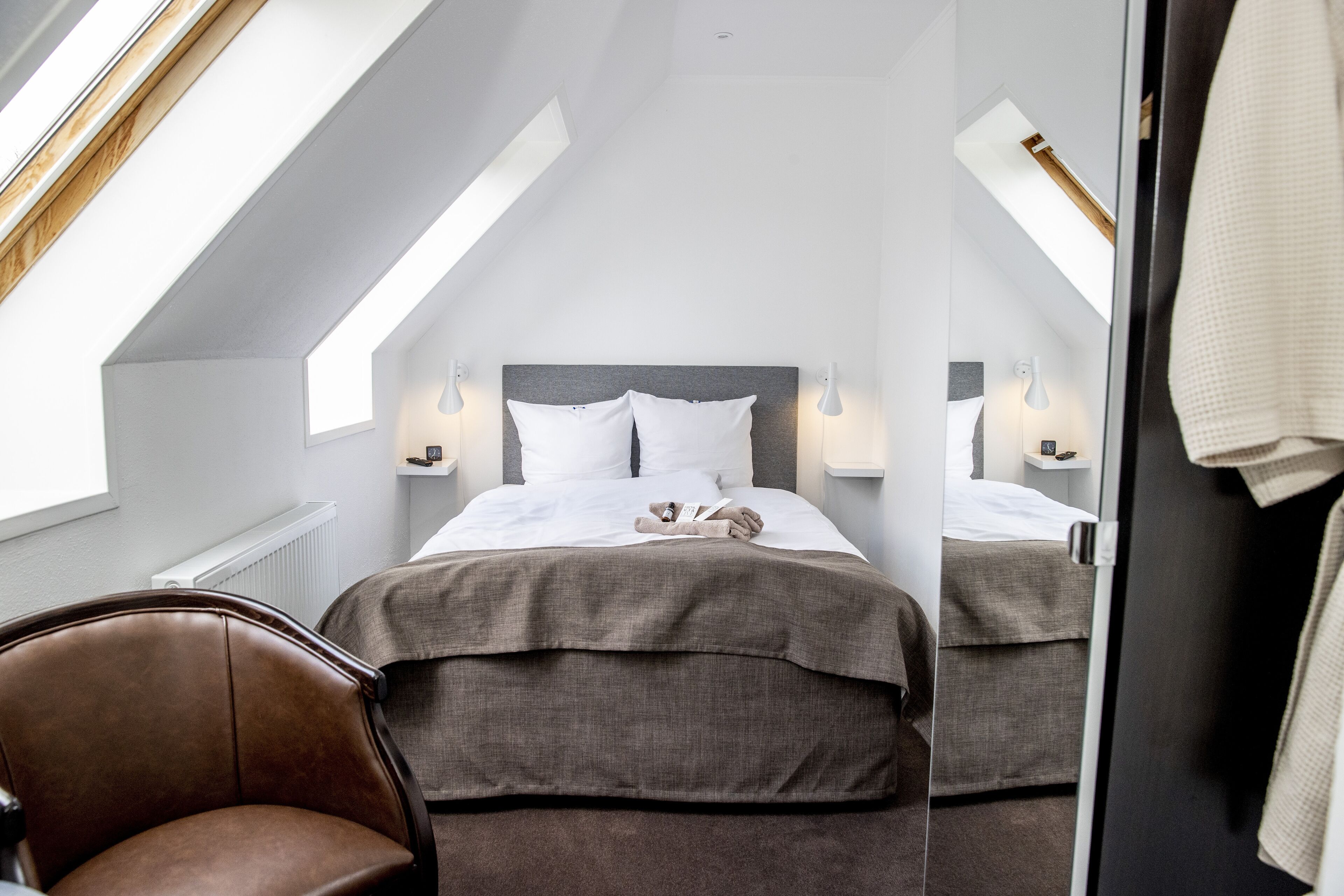 Um quarto moderno com teto inclinado, uma cama grande com cabeceira cinza, luminárias laterais e uma poltrona de couro marrom.