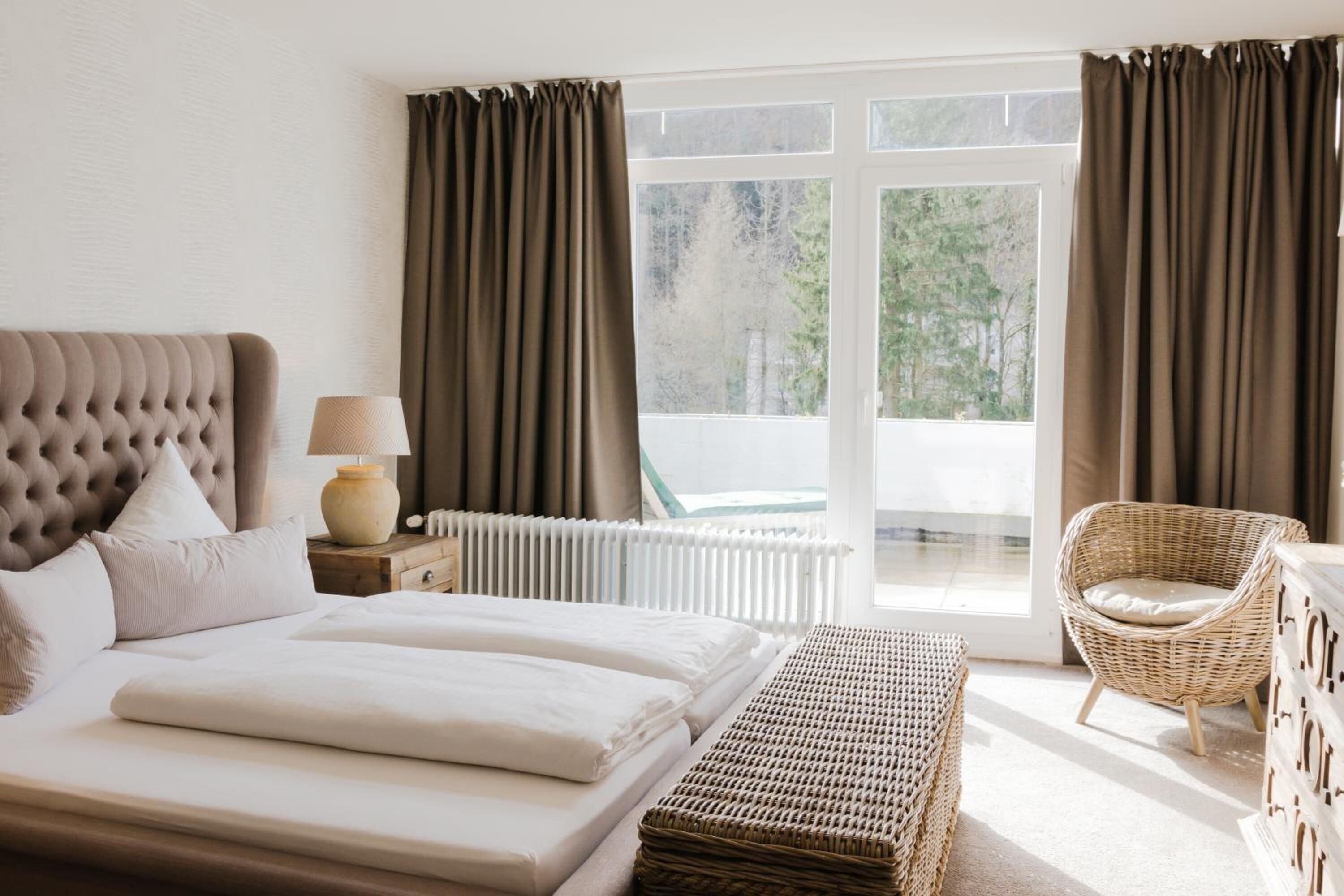 Quarto com uma cama grande, mesa de cabeceira com abajur, cadeira de vime e vista para o exterior através de uma janela ampla.