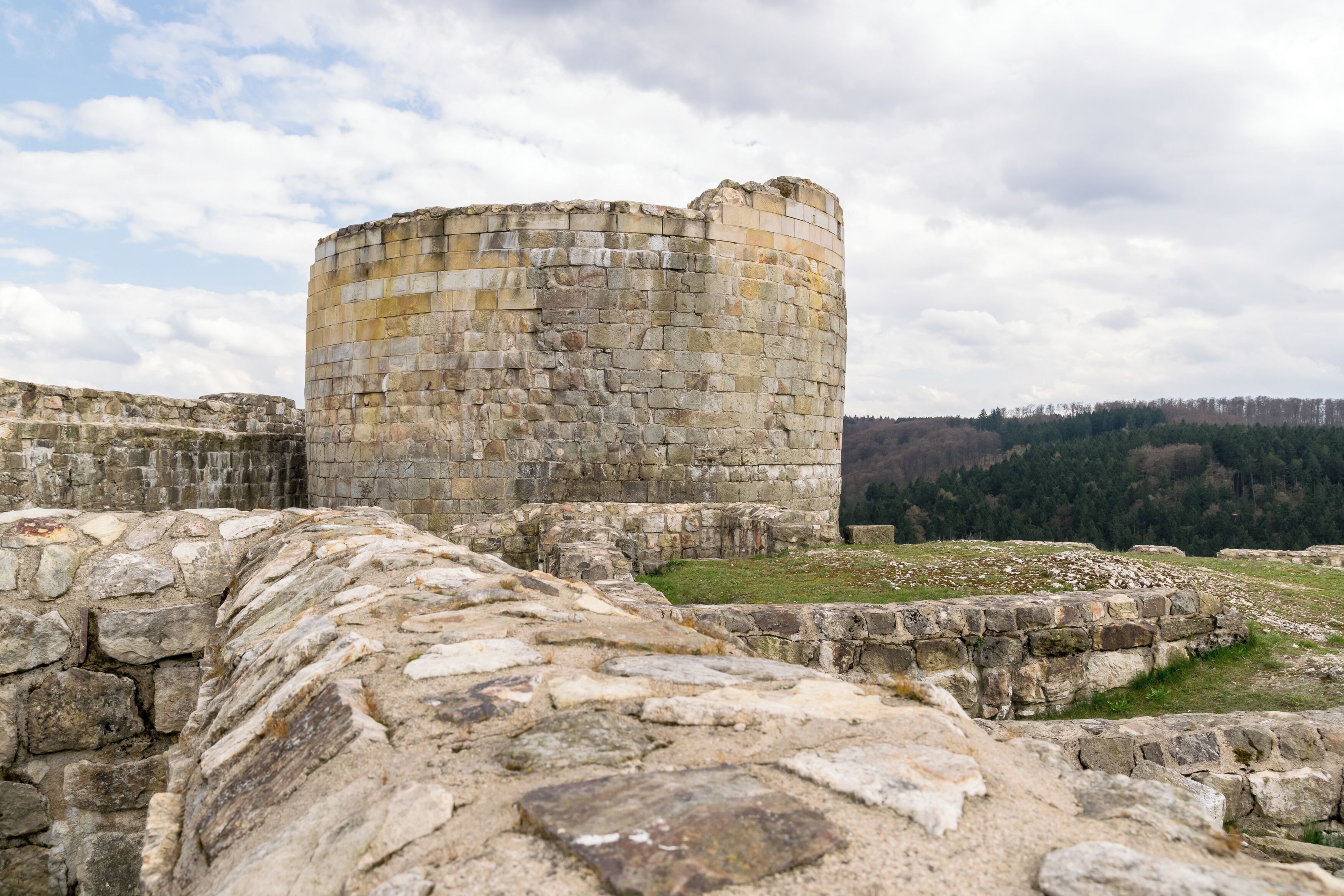 Ruínas de um castelo de pedra com uma torre circular, cercado por muros de pedra e uma área gramada.