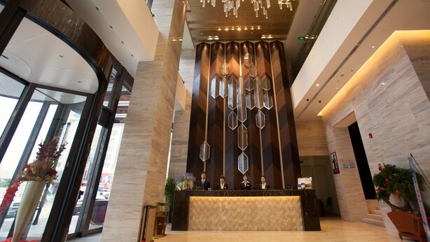 Um hall de hotel moderno com uma mesa de recepção, grandes janelas e um lustre decorativo no teto.
