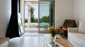 Uma sala de estar moderna com um sofá, uma mesa de centro com um livro e um vaso de flores, e uma porta de vidro deslizante que dá para uma área externa com piscina e vegetação.