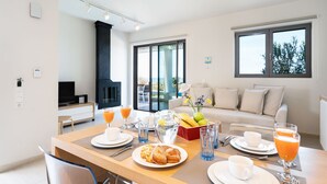 Uma zona de refeições moderna com uma mesa de madeira posta para o pequeno-almoço, incluindo pratos de pastelaria, uma taça de fruta e copos de sumo de laranja.