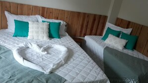 Quarto de hotel com duas camas, cada uma com roupa de cama branca e almofadas verde-água. Possui cabeceira de madeira e um banquinho de madeira.