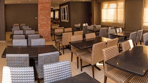 Uma área de refeições de hotel com várias mesas e cadeiras, um pilar de tijolos e grandes janelas com cortinas.