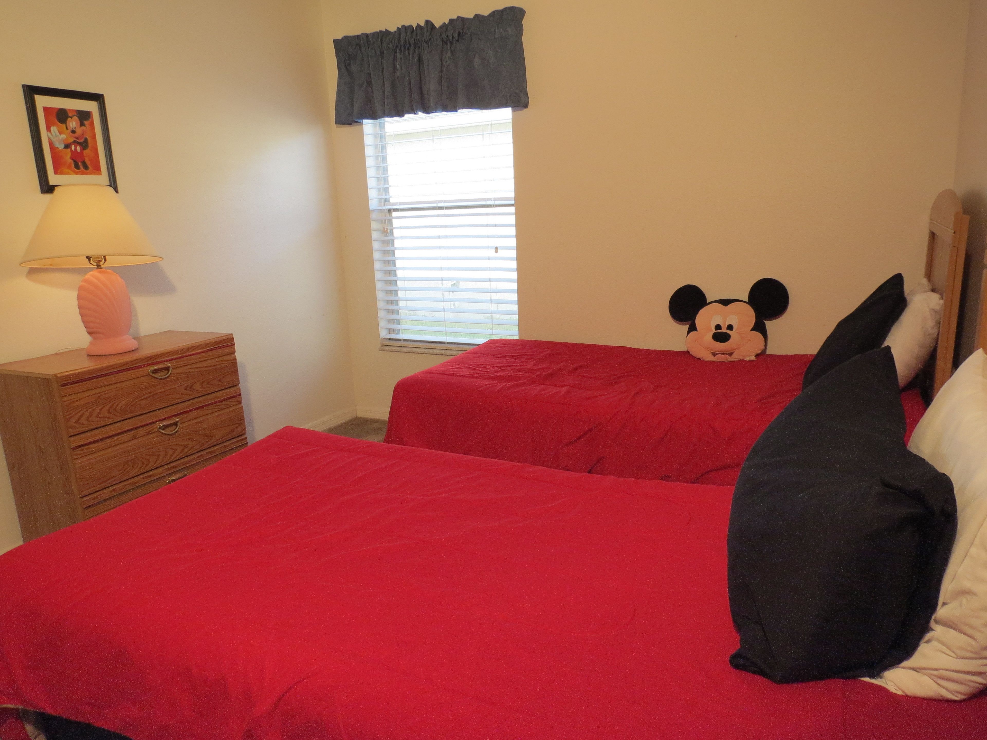 Quarto com duas camas, uma mesa de cabeceira com abajur, uma janela com persianas e uma almofada com o rosto do Mickey Mouse em uma das camas.