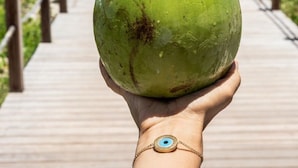 Uma mão segurando um coco verde com um canudo, em um calçadão de madeira que leva a uma praia.