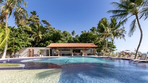 Piscina transparente com espreguiçadeiras e área de estar coberta, rodeada por palmeiras.