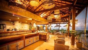 Uma área de bar com balcão de madeira, móveis de vime e vista para o oceano.