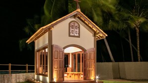 Uma pequena construção de madeira bem iluminada com uma cruz no topo, possivelmente uma capela ou igreja, cercada por um muro de pedra e palmeiras.