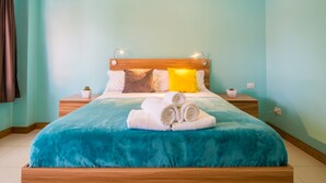 Uma cama bem arrumada com um cobertor azul, duas mesas de cabeceira com abajures, e um pequeno vaso de planta em cada mesa.