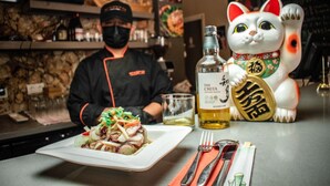 Um chef preparando um prato em restaurante, com uma estátua decorativa de gato e uma garrafa de bebida na bancada.