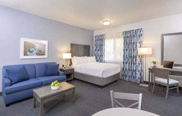 Quarto de hotel com cama, sofá azul, escrivaninha com cadeira, mesinha com frutas e luminária.