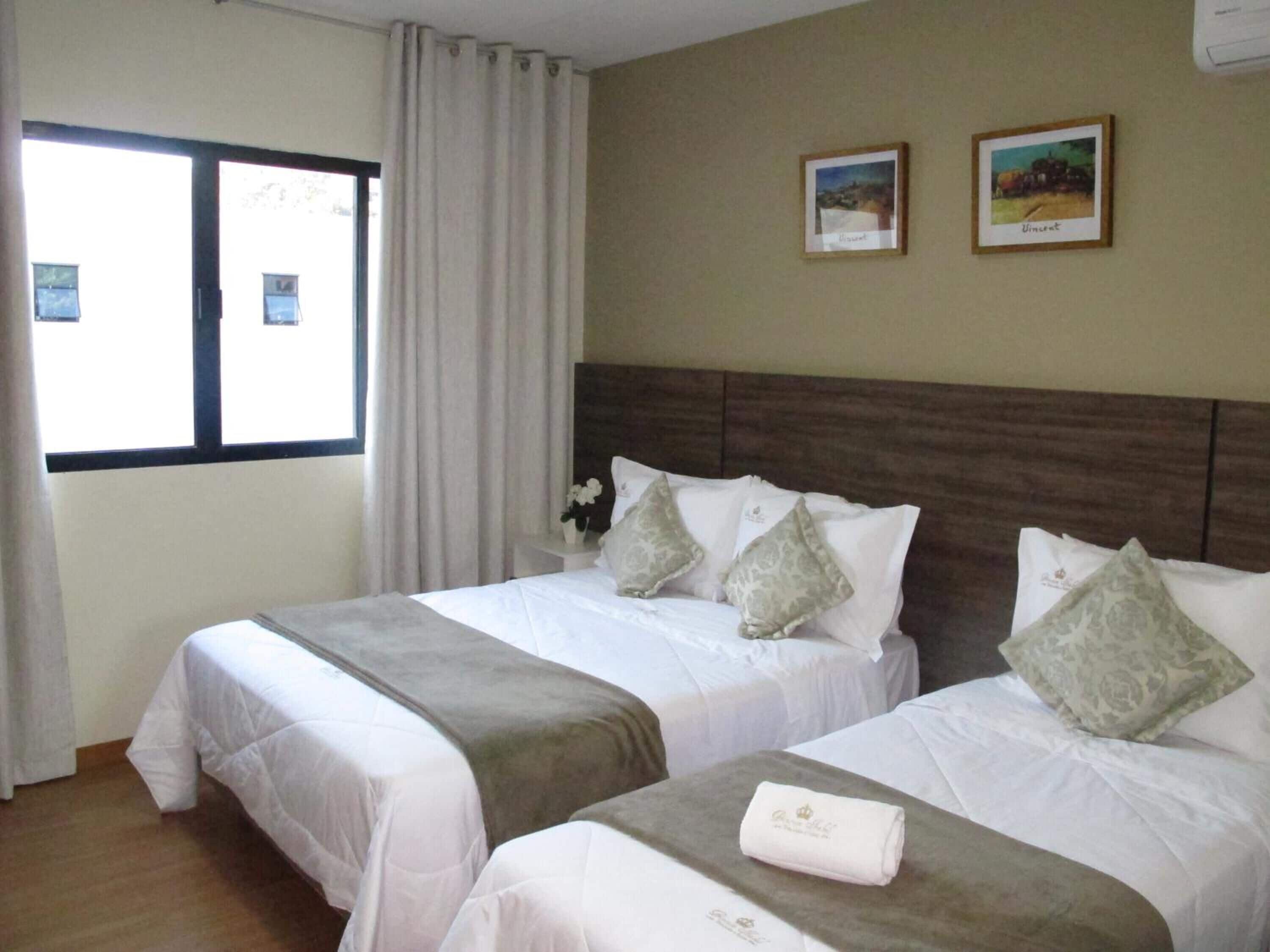 Quarto de hotel com duas camas, uma janela grande e duas imagens emolduradas na parede.