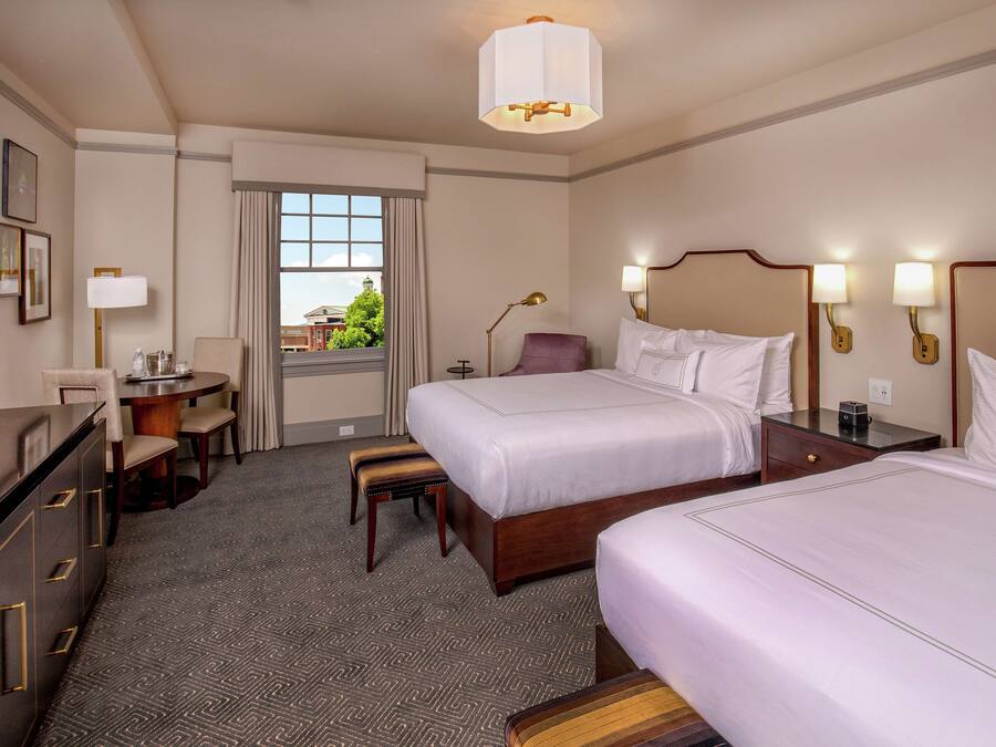 Quarto de hotel com duas camas, uma mesa, uma cadeira, uma luminária e vista para o exterior.