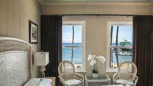 Quarto de hotel com uma cama, duas cadeiras de vime, uma mesinha com abajur e vista para o mar e barcos através de duas janelas.