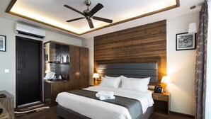 Quarto de hotel com cama grande, cabeceira de madeira, luminárias laterais, ventilador de teto e guarda-roupa.